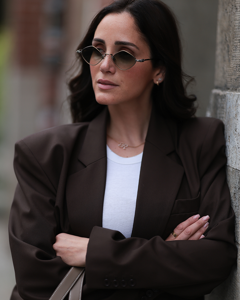 Close-up of brown blazer and white ribbed top styled with tinted oval sunglasses and delicate jewelry outdoors