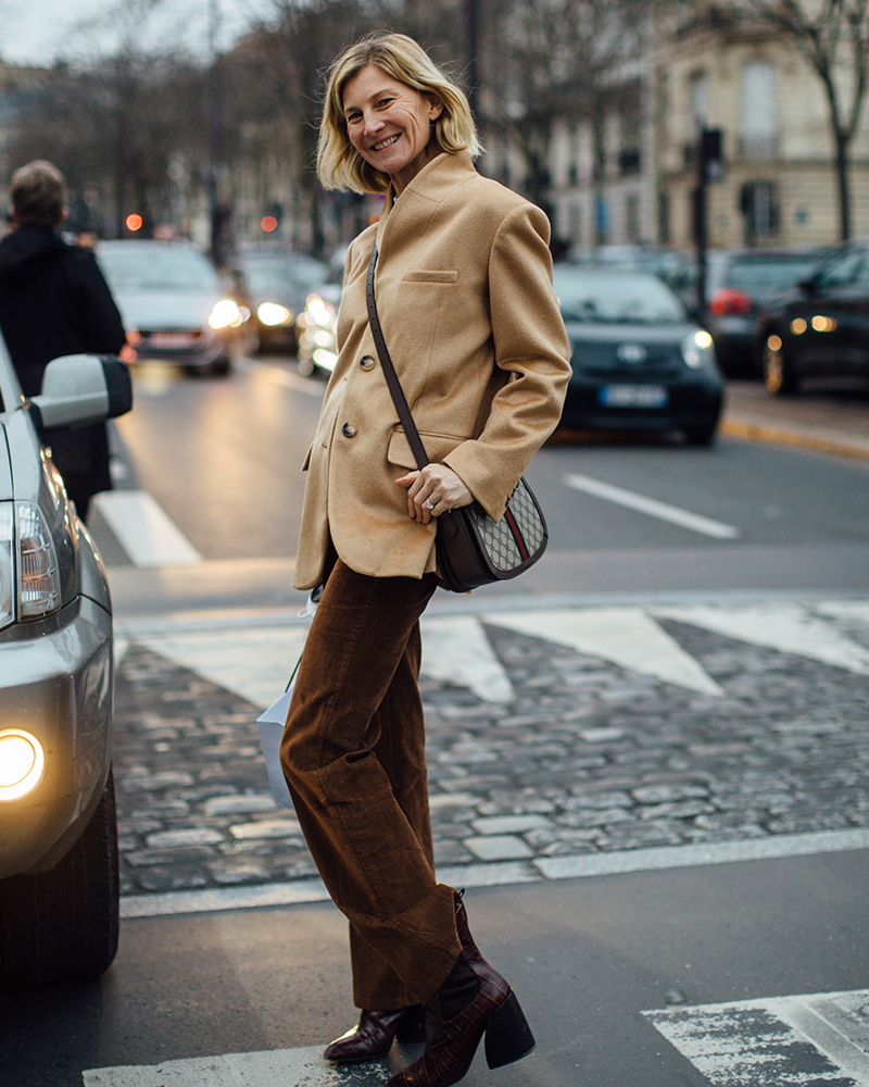 Model in brown corduroy pants%2C camel blazer and leather ankle boots with a crossbody bag on a city street