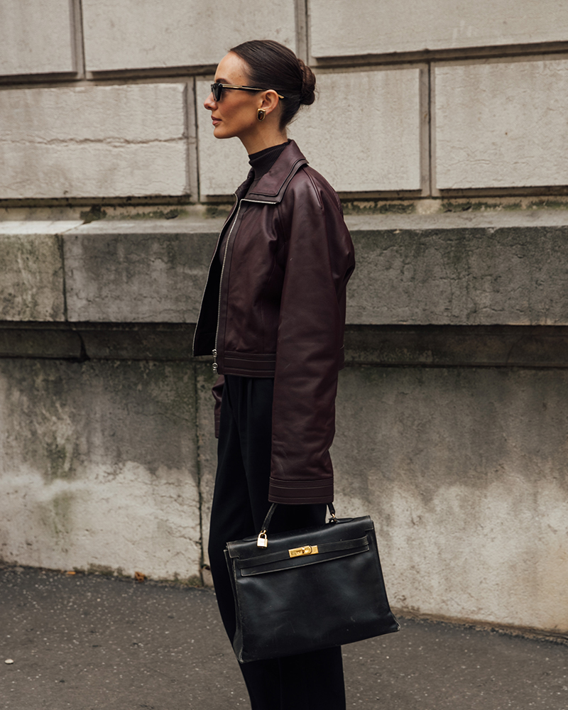 Street style look featuring a burgundy leather jacket%2C black trousers and structured handbag against a stone wall