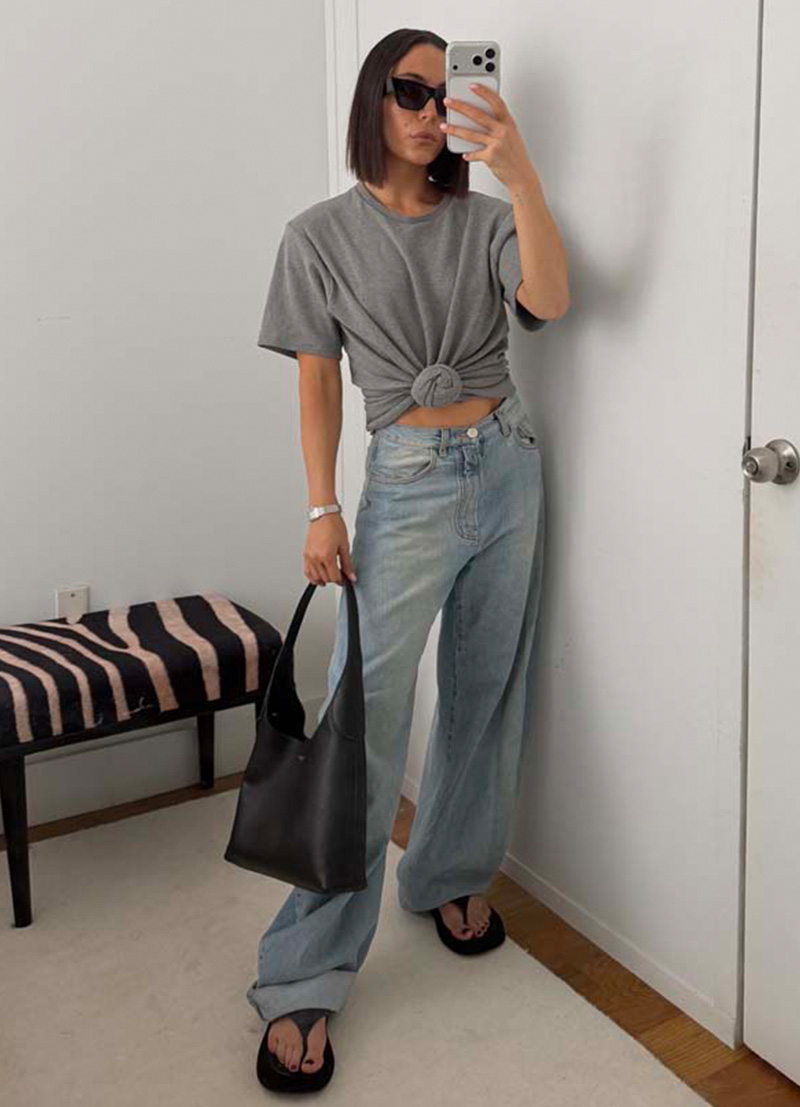 Mirror selfie in a knotted gray T-shirt, wide-leg jeans, black tote bag and sunglasses in a minimalist room