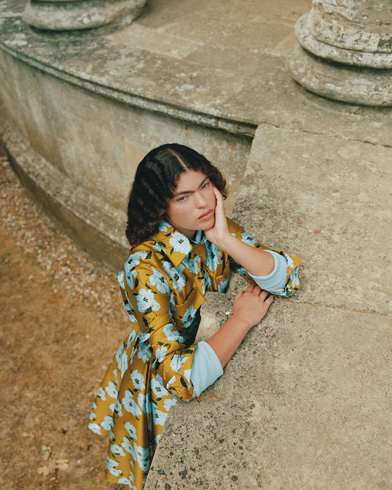 Mustard floral midi dress with blue blossoms and bow neckline%2C posed beside weathered stone architecture