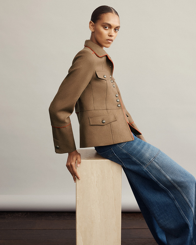 Model in a brown military-style jacket with red piping and wide-leg blue jeans%2C posed in a minimalist studio%0A