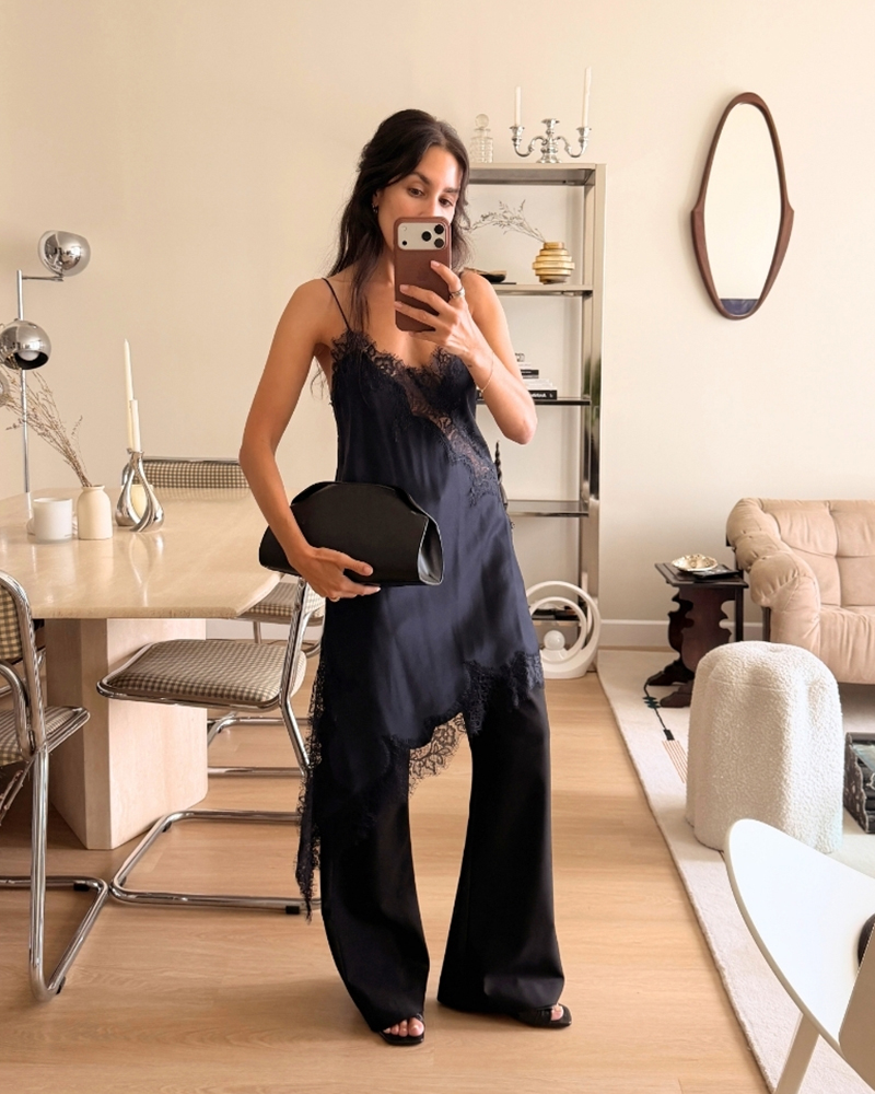 Woman wearing black silk lace-trimmed slip dress layered over wide-leg pants%2C holding black clutch%2C styled for pajama trend indoors