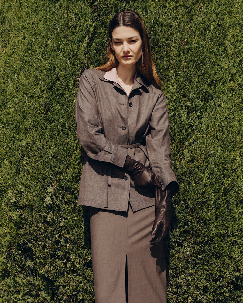 Brown belted tailored jacket and matching midi skirt styled with long leather gloves against a lush green hedge%0A
