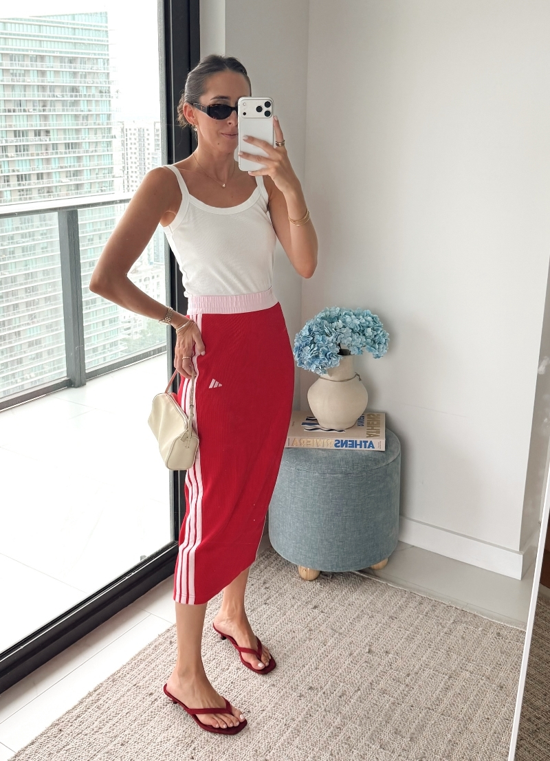 White tank top with red Adidas midi skirt, cream shoulder bag and flip-flops in a modern apartment