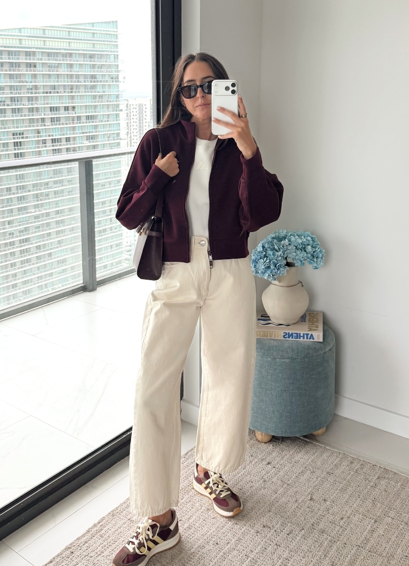Model in a burgundy Adidas jacket and grey flared pants, carrying a suede tote beside black luggage indoors