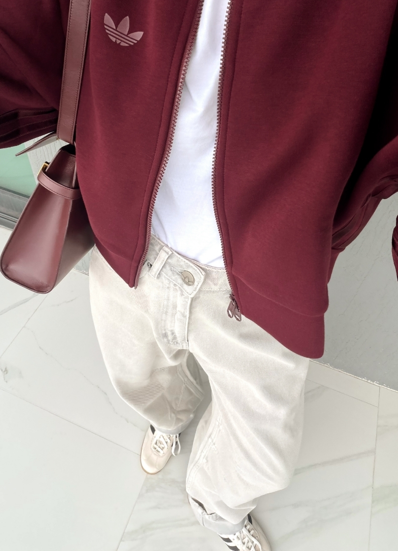 Close-up of burgundy Adidas zip jacket, cream jeans and leather shoulder bag styled with sneakers indoors