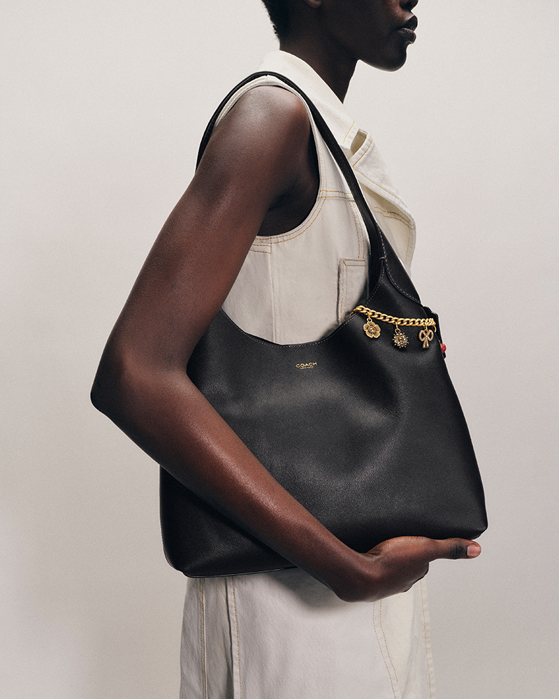 Model carrying a black Coach leather tote with gold charms%2C styled with an ivory sleeveless dress in a studio