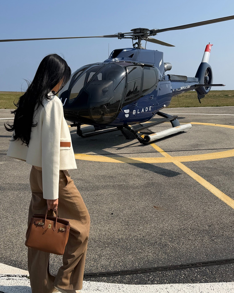 Cream cropped jacket with tan trousers and a brown top-handle bag styled beside a Blade helicopter on a helipad
