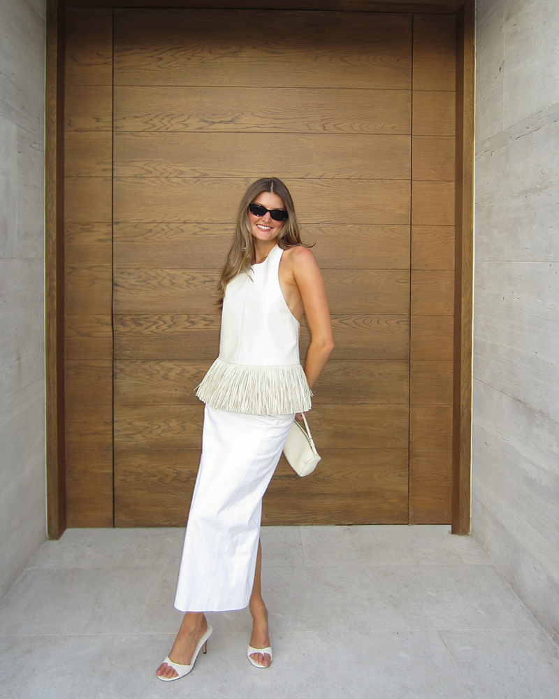White party outfit featuring a fringed sleeveless top%2C midi skirt%2C sunglasses and heels against a wood-paneled backdrop