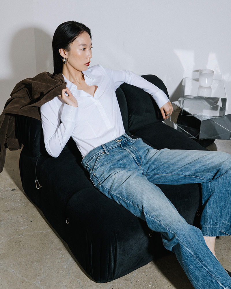 White button-down shirt styled with relaxed blue jeans and gold hoop earrings on a black lounge chair%0A