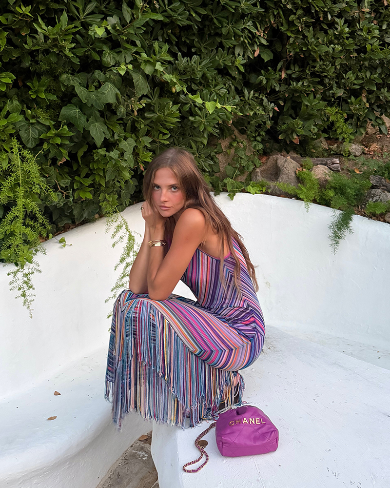 Model in a colorful striped fringe maxi dress with a pink Chanel bag%2C seated on a white garden bench