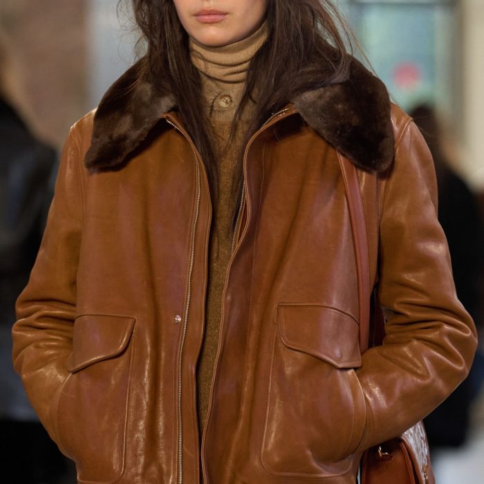 Brown leather bomber jacket with dark fur collar, paired with tan turtleneck and matching leather bag for fall fashion