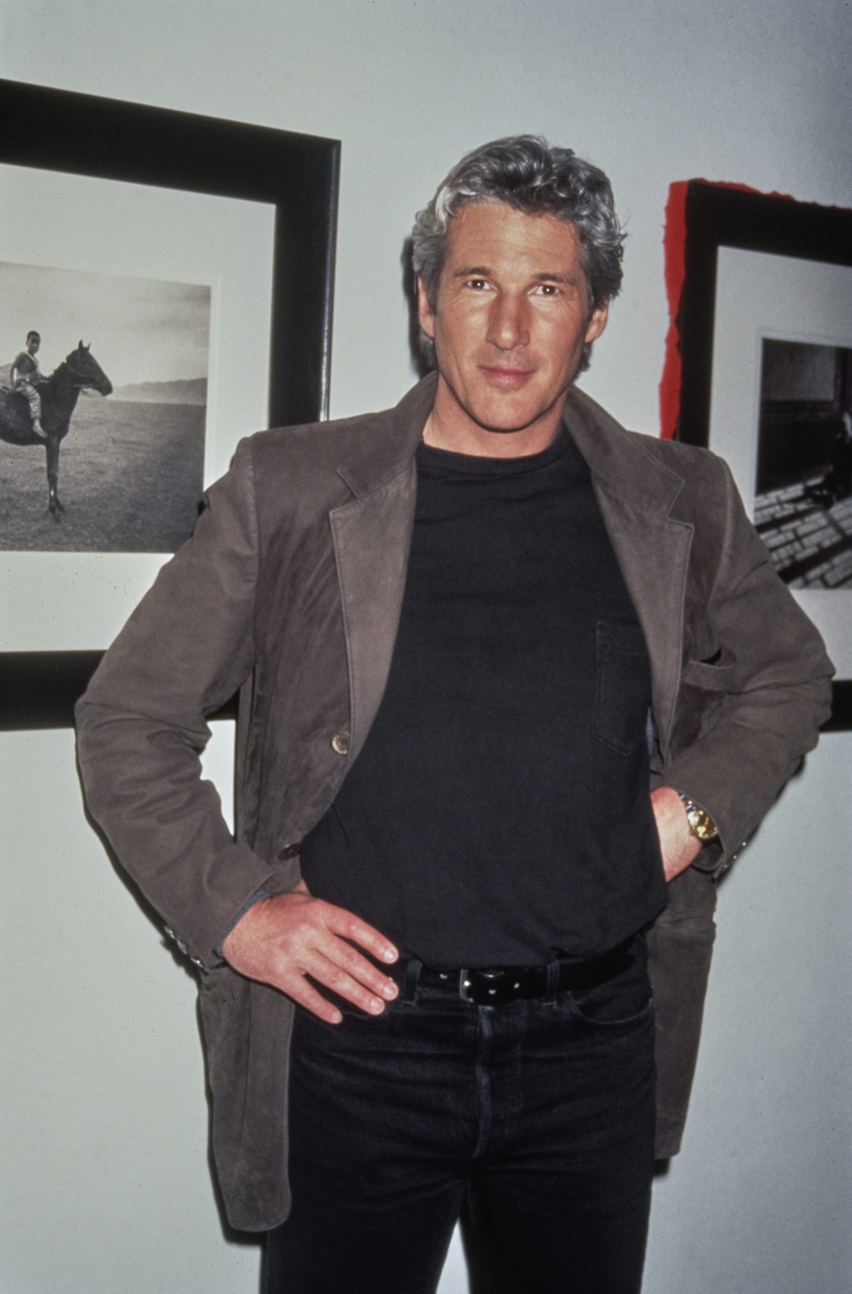 Man wearing a brown suede blazer over a black crew neck t-shirt with black jeans and a black leather belt in an indoor gallery setting
