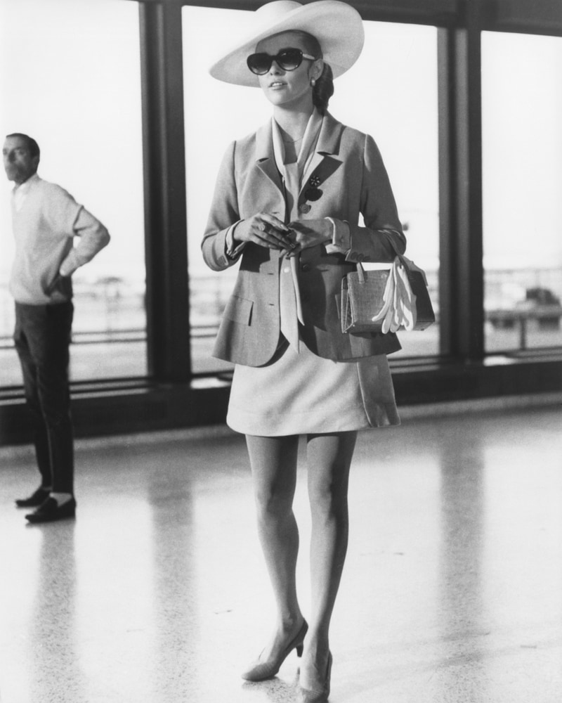Woman wearing a tailored blazer and mini skirt with a wide-brimmed hat%2C oversized sunglasses%2C and carrying a structured handbag with gloves%2C showcasing classic 1960s fashion style