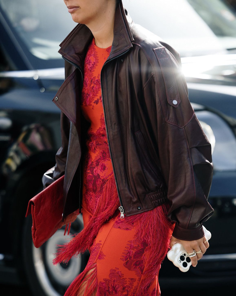 Woman wearing a red fringe midi dress with floral patterns paired with a dark brown leather bomber jacket and a red clutch bag