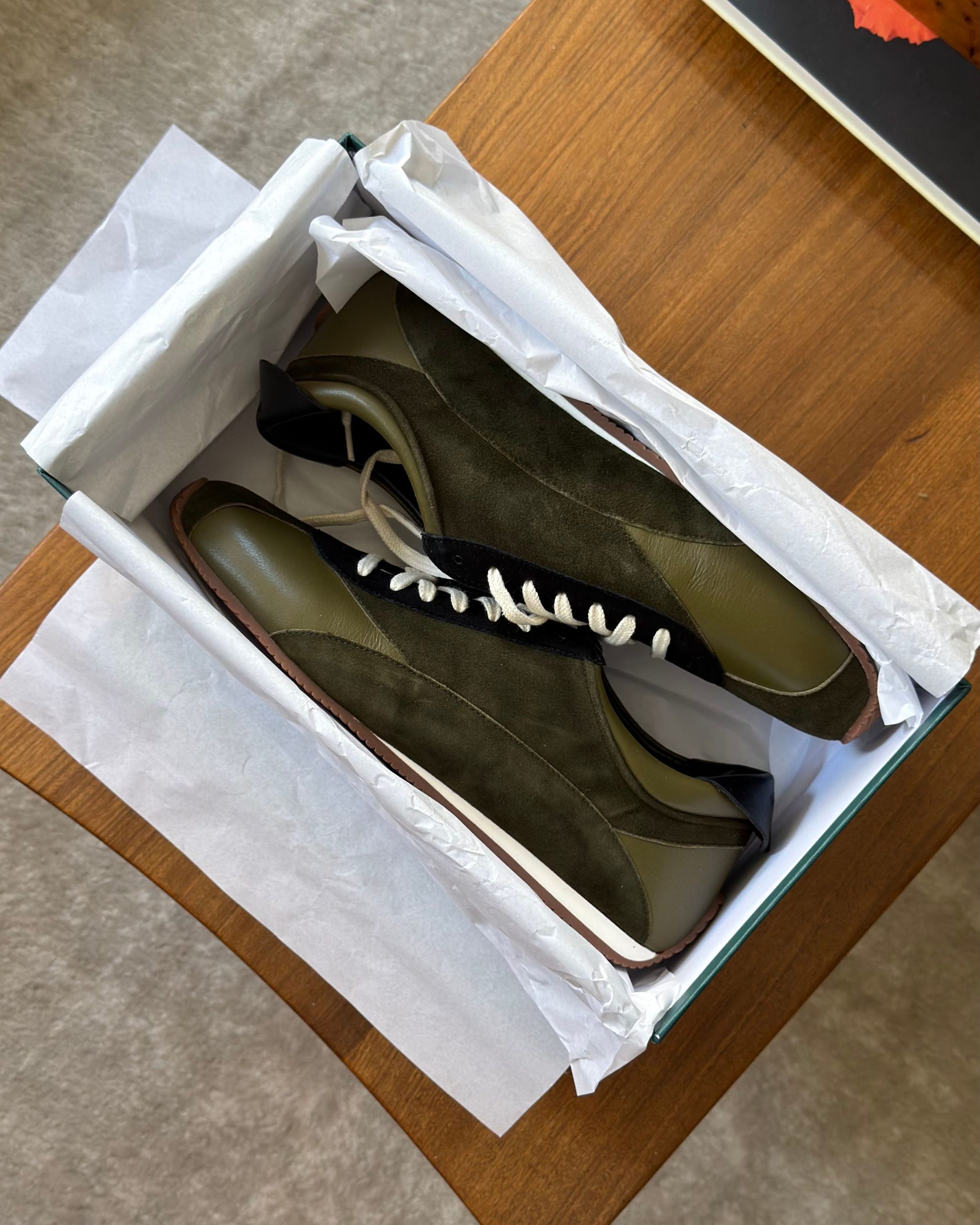 Olive green suede and leather sneakers with white laces in a shoebox on a wooden table