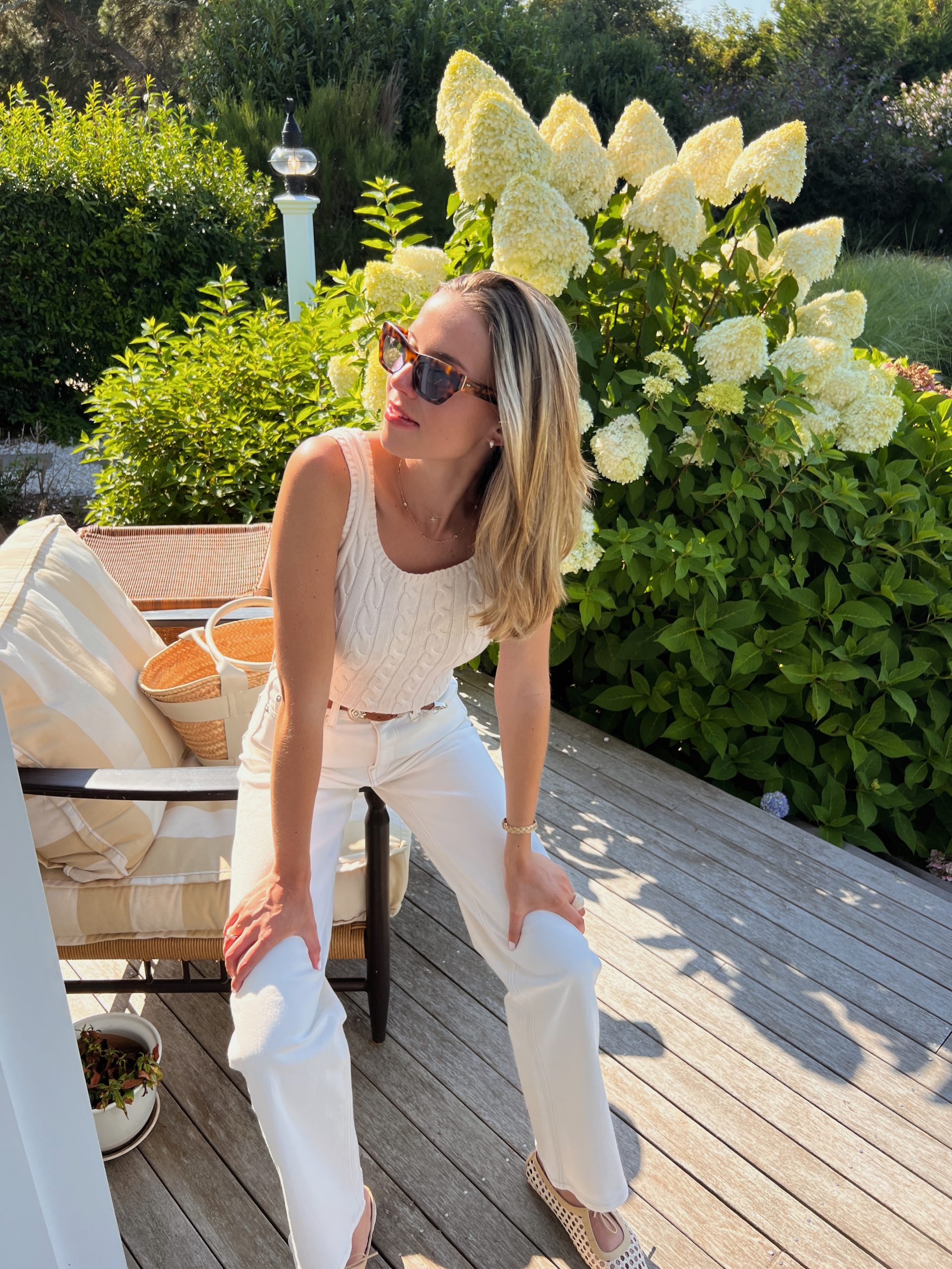 Woman wearing a sleeveless white cable knit crop top with high-waisted white pants and tortoiseshell sunglasses in a sunny garden setting