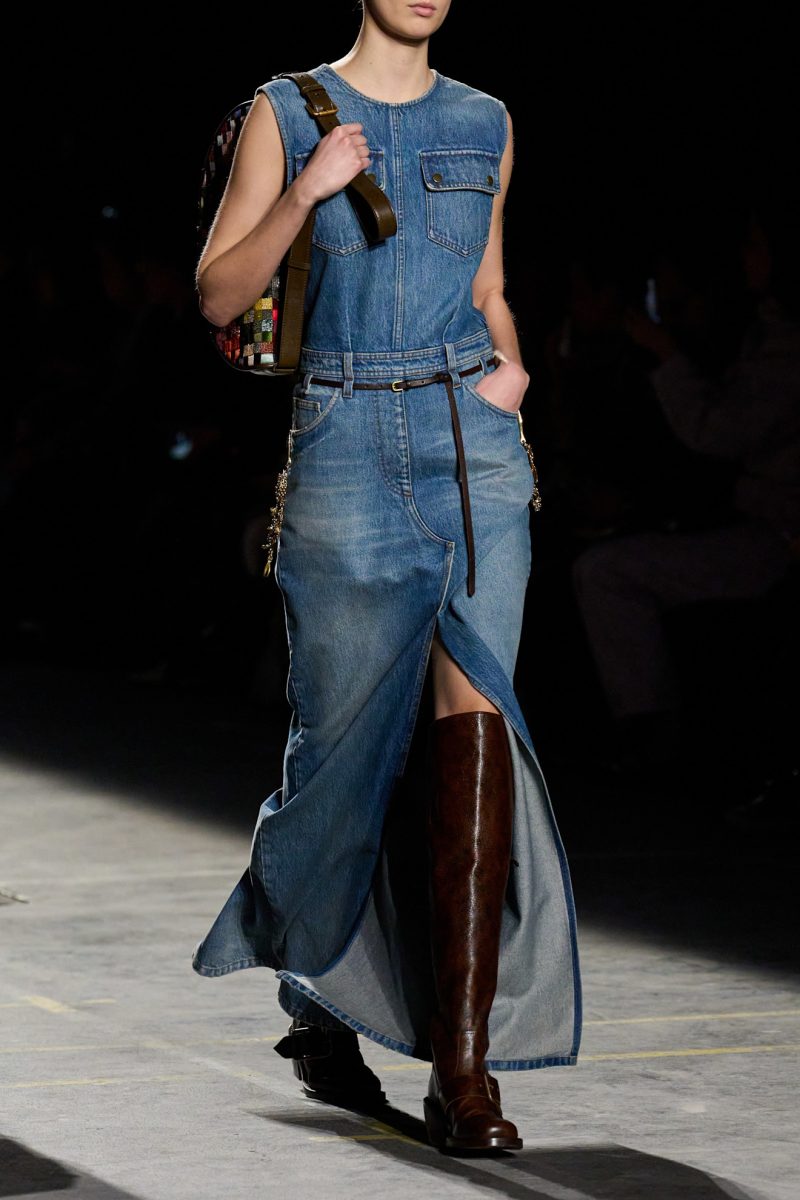 Model wearing sleeveless denim maxi dress with front slit, brown leather knee-high boots, and a patterned backpack on runway