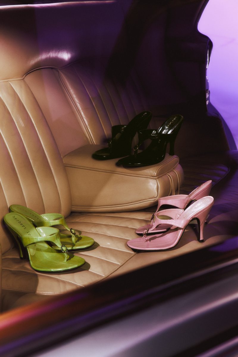 Three pairs of heeled sandals in black, pink, and green patent leather displayed on a vintage tan leather car seat, showcasing retro summer footwear style