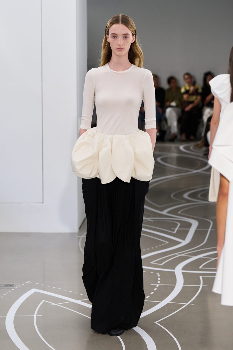 Model wearing a cream fitted top with three-quarter sleeves and a sculptural cream peplum detail paired with a flowing black maxi skirt on runway