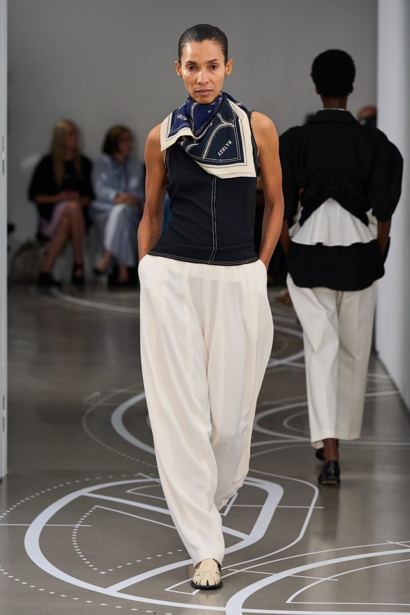 Model wearing a sleeveless black top with white stitching, loose white trousers, cream woven shoes, and a navy blue Ashlyn silk scarf styled around the neck