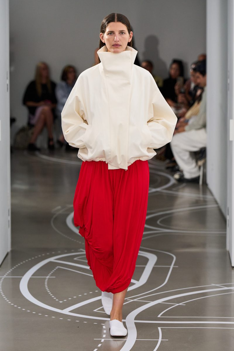 Model wearing a cream oversized high-collar jacket paired with flowing red wide-leg pants and white slip-on shoes on runway