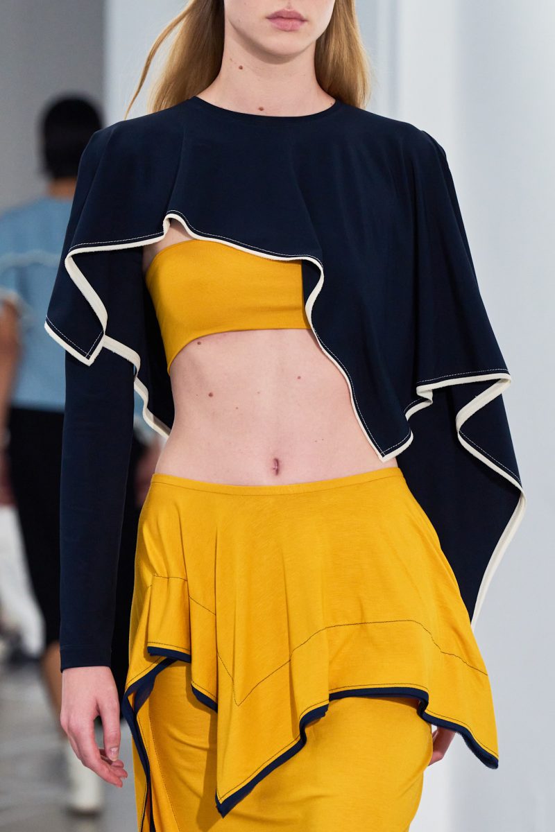 Navy cropped jacket with white trim over mustard yellow bandeau top and asymmetrical skirt with navy edging in a modern layered outfit