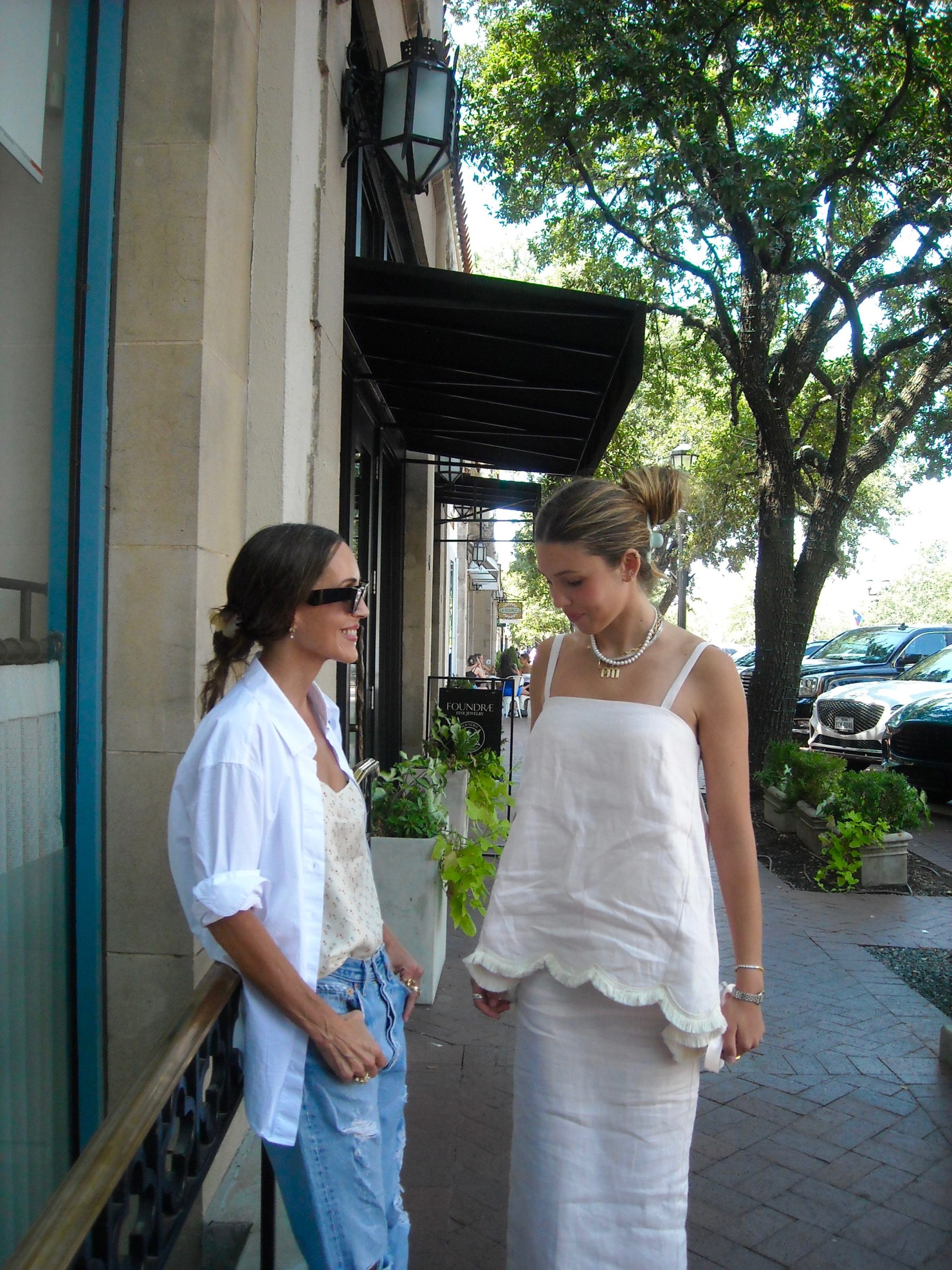 Two women in casual summer outfits%2C one wearing a white linen tank top and skirt with pearl necklace%2C the other in a white shirt over a floral top and ripped jeans