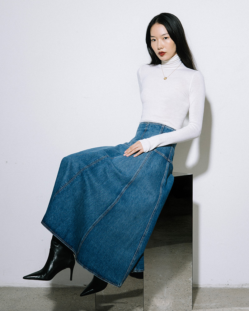 White turtleneck paired with a long blue denim skirt and black pointed boots in a minimalist studio setting