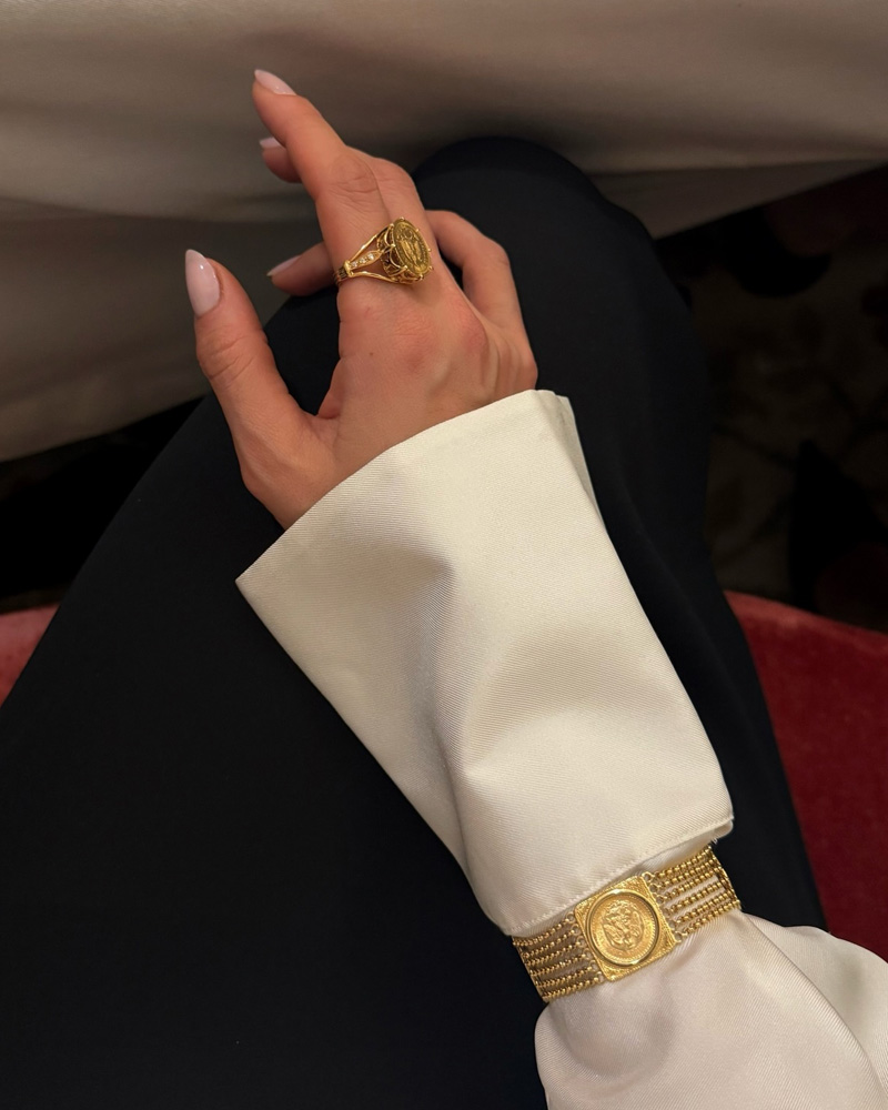 Close-up of gold signet ring and medallion bracelet styled with a white cuff and black trousers