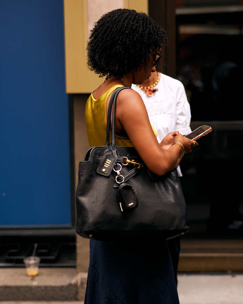 Black leather tote bag with gold hardware styled with a yellow satin top and navy skirt on a city street%0A