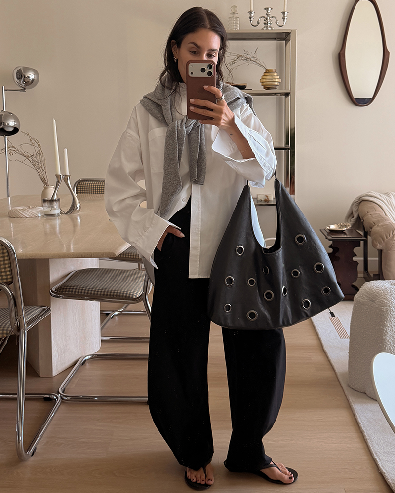 Model in an oversized white shirt and black trousers carrying an eyelet leather tote%2C taking a mirror selfie at home