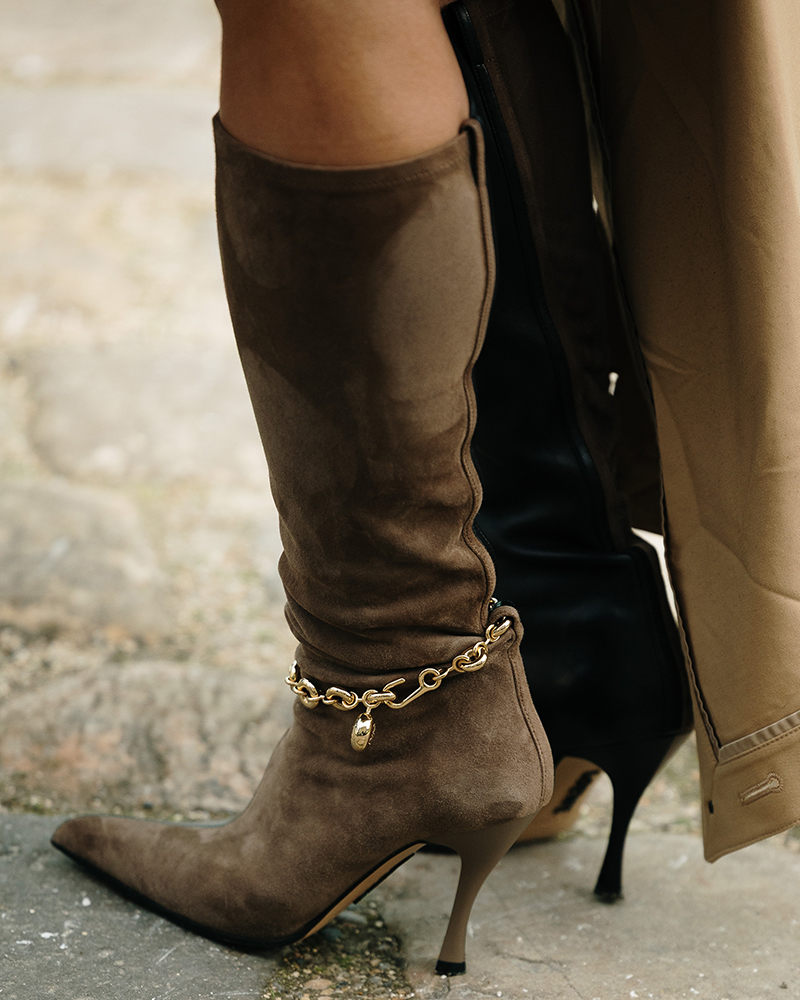 Brown suede knee-high boots with pointed toes and gold chain ankle detail%2C styled on a stone pavement