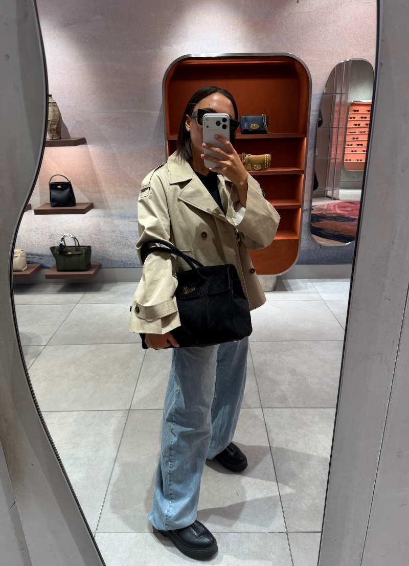 Beige cropped trench jacket with light-wash jeans and black suede tote, photographed in a boutique mirror