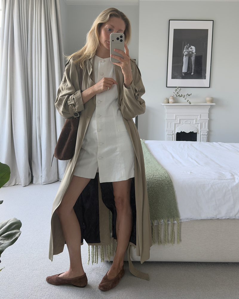 Ivory linen mini dress layered under a taupe trench coat with brown flats and suede bag in a serene bedroom