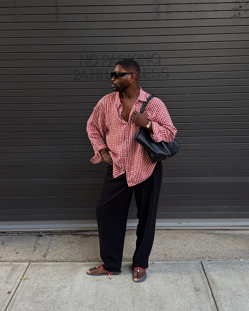 Man in a red gingham shirt and black trousers with a leather shoulder bag and sunglasses%2C styled on a city sidewalk