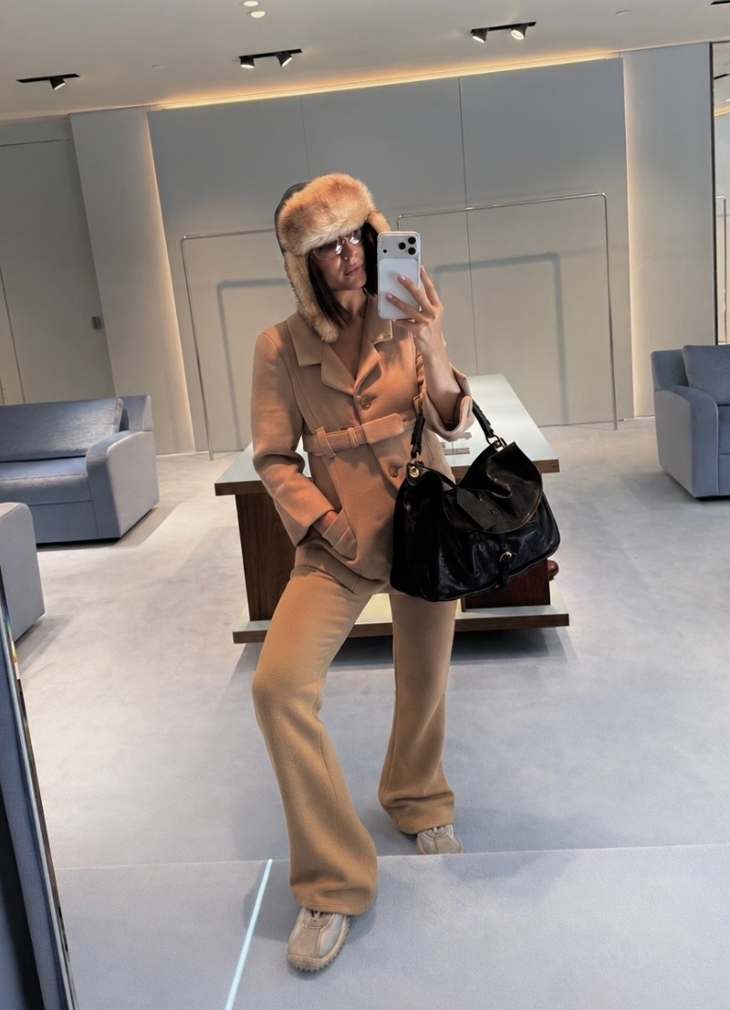 Camel belted suit styled with a fur trapper hat, oversized black leather bag and cream sneakers in a boutique