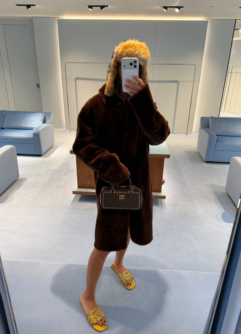 Brown faux-fur coat styled with a fur trapper hat, studded Miu Miu bag and yellow embellished slides in store