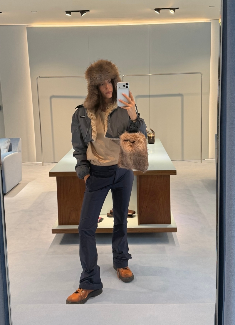 Sporty layered look with fur trapper hat, gray jacket, navy trousers, fur bag and brown shoes in a boutique