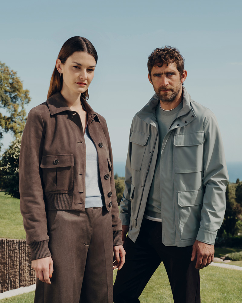 Models wearing brown and blue jacket against a lush green hedge