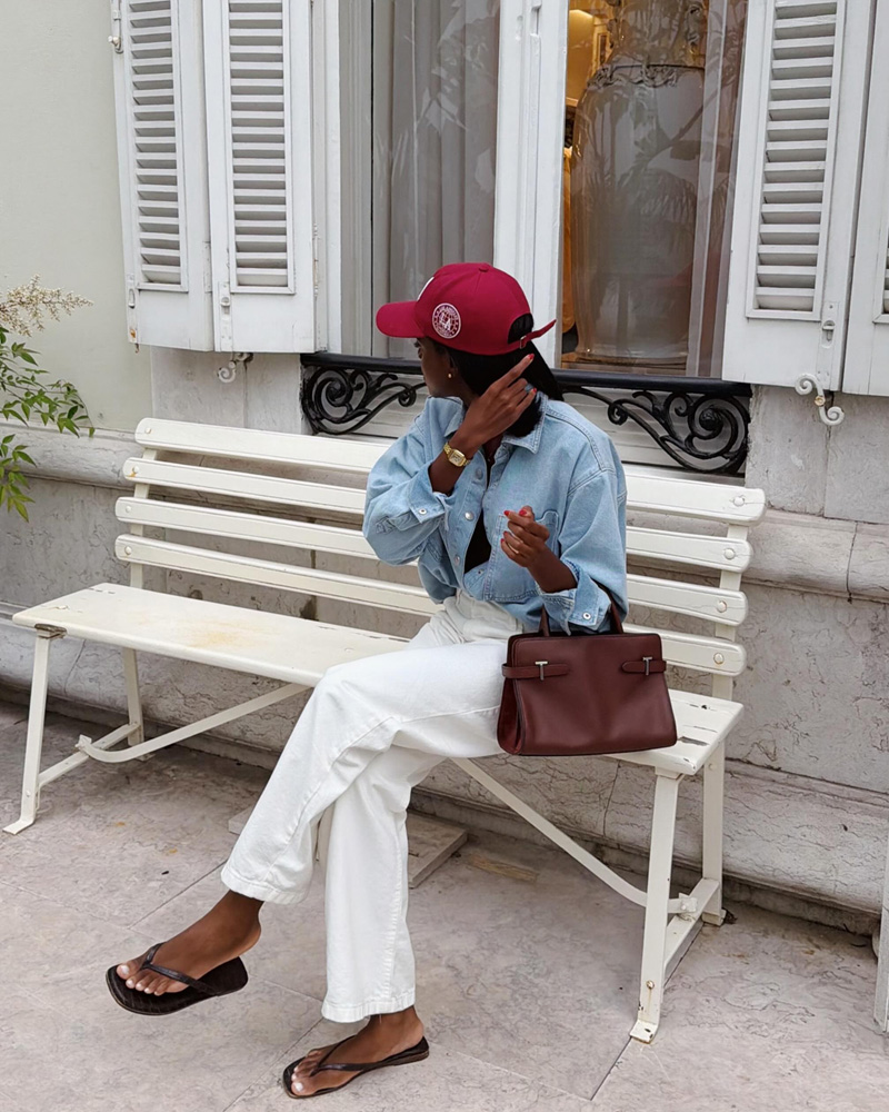 Relaxed denim shirt styled with white jeans%2C burgundy cap and structured leather handbag on an outdoor bench%0A