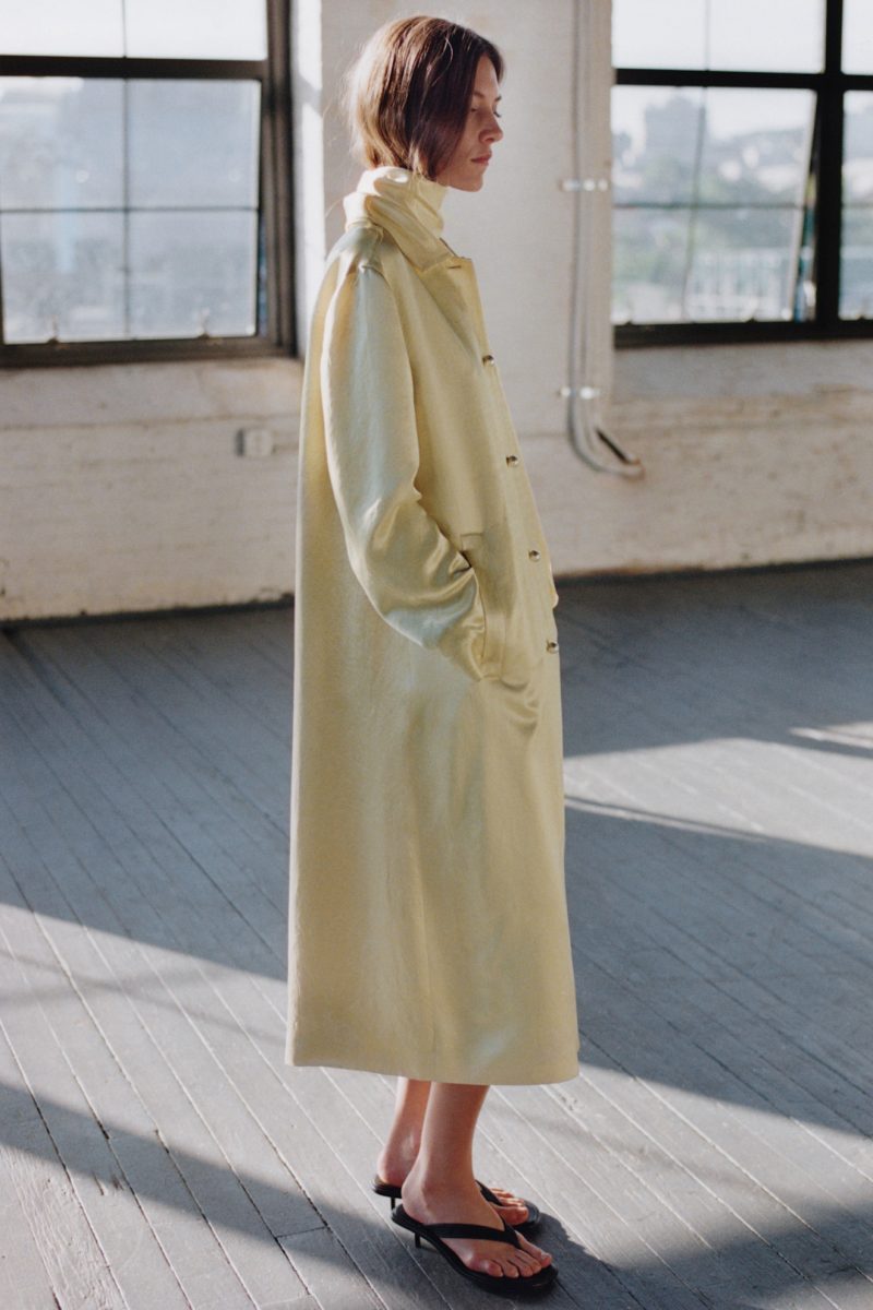 Model wearing a long pale yellow wool coat with button details and pockets paired with black heeled sandals in a sunlit industrial space