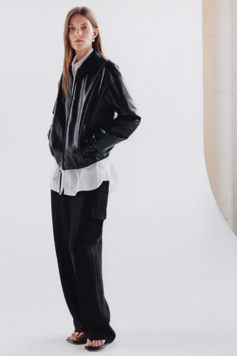 Model wearing a black leather jacket over a white oversized shirt, paired with loose black cargo pants and black sandals in a minimalist studio setting
