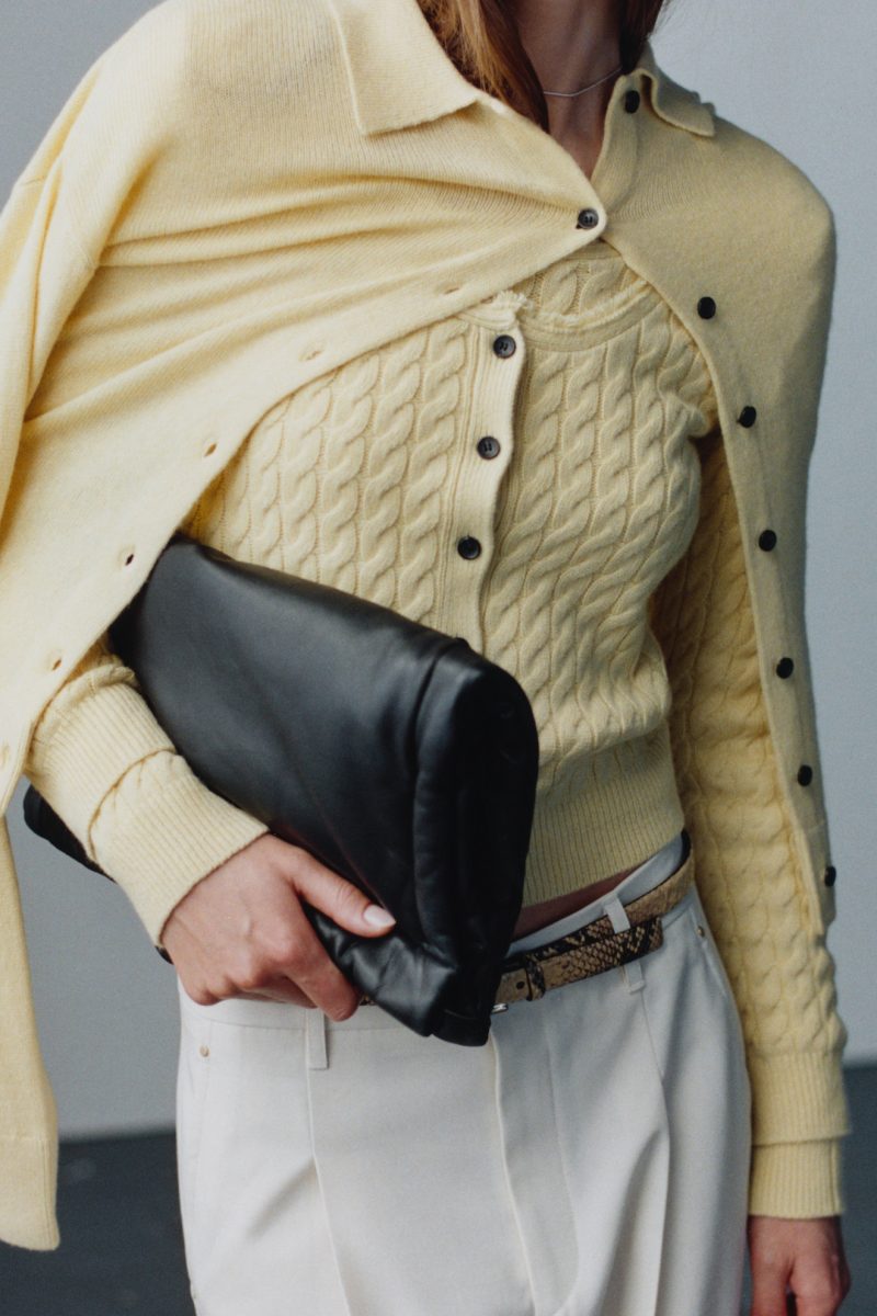 Yellow cable knit cardigan and matching sweater layered over white tailored trousers with a snakeskin belt and black leather clutch bag