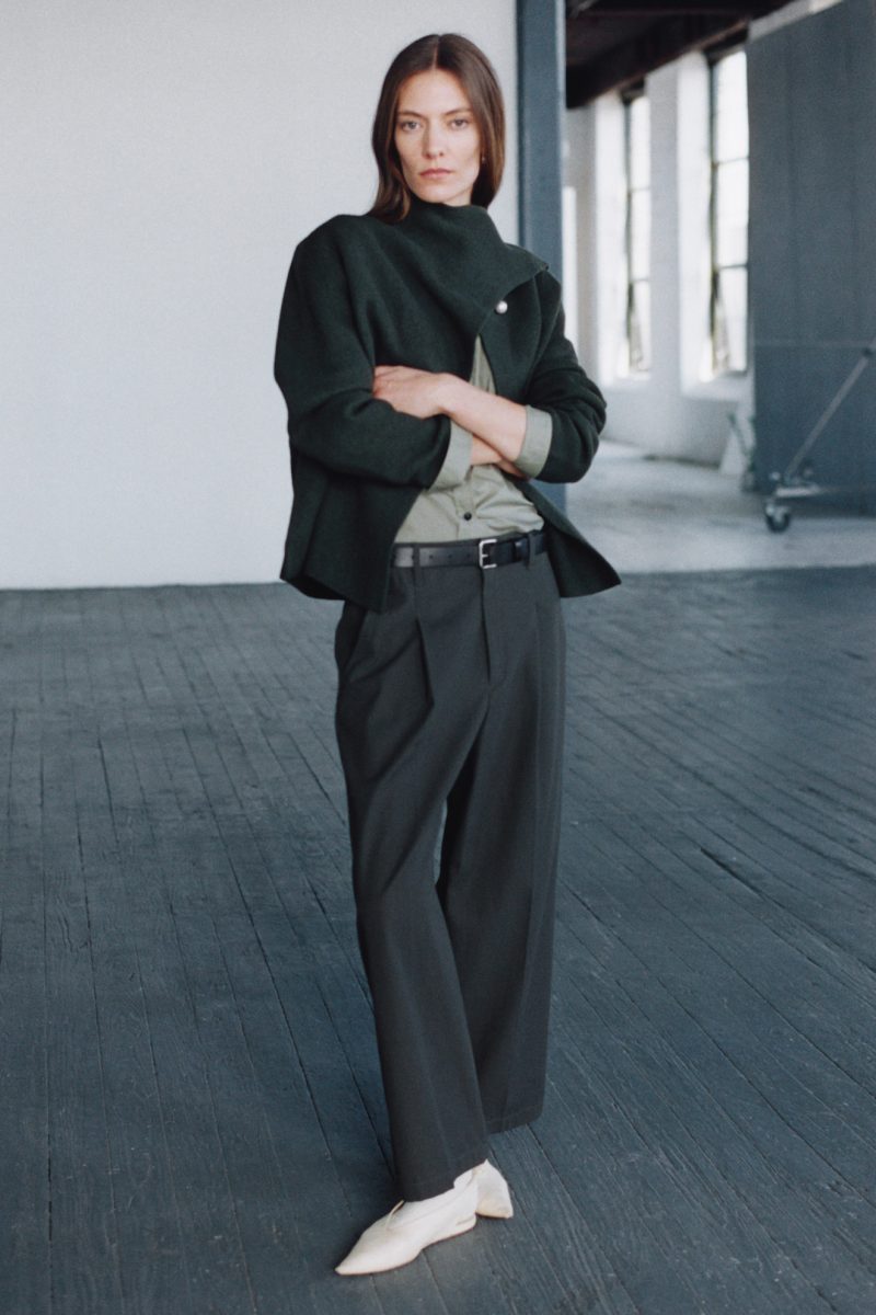 Model wearing a dark green asymmetrical wool jacket over a light olive button-up shirt, paired with high-waisted charcoal wide-leg trousers and white pointed flats in a minimalist indoor setting