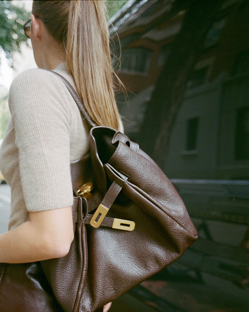Brown leather slouchy tote bag with modern hardware details%2C paired with a light knit top for a casual%2C chic look in an urban setting