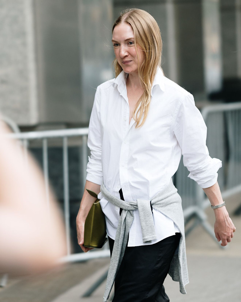 Woman wearing a white button-up shirt with a gray sweater tied at the waist%2C black midi skirt%2C and carrying an olive green clutch bag