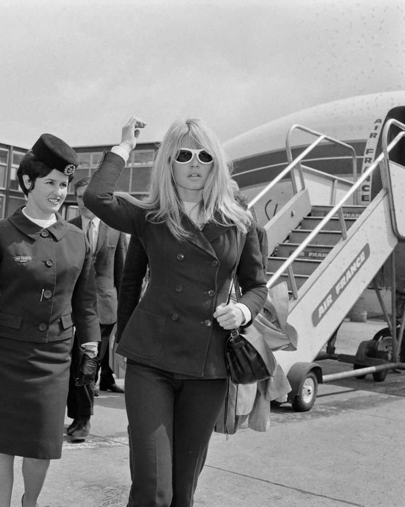 Brigitte Bardot exudes vintage glamour in a tailored black double-breasted coat and chic sunglasses%2C strolling through the airport with confidence.