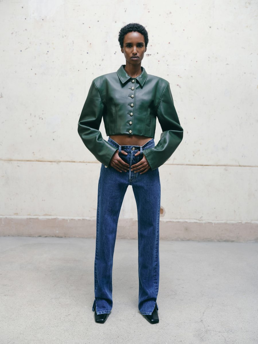 Model wearing a cropped dark green leather jacket with silver buttons paired with high-waisted straight-leg blue jeans and black pointed shoes
